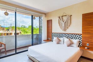 Quarto de hotel com uma cama grande, cabeceira de madeira, um painel de macramé na parede e vista para um canteiro de obras através de uma porta de vidro.