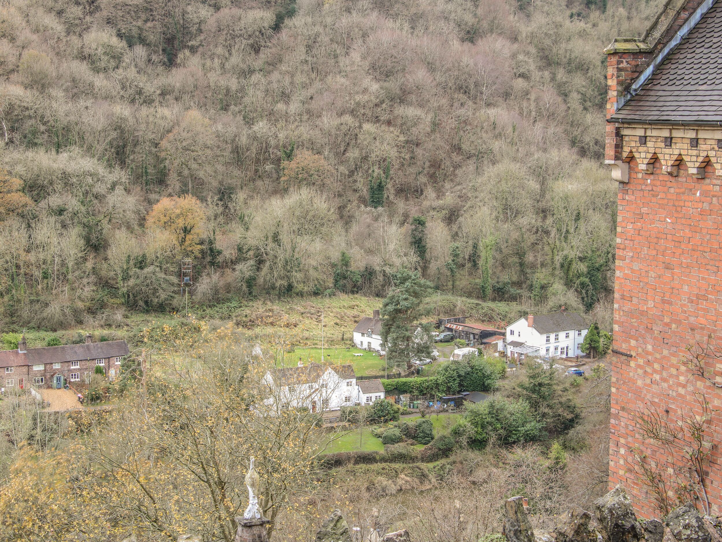 Uma vila rural com casas tradicionais, um edifício de tijolos em destaque e colinas arborizadas ao redor.