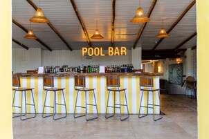 Um bar à beira da piscina, com balcão de madeira, banquetas de metal e luminárias de vime penduradas.