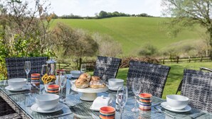 Mesa de jantar ao ar livre posta com pratos, tigelas e copos listrados brancos, com vista para um campo verde e árvores ao fundo.