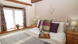 Quarto com cama grande, cabeceira de madeira, duas mesas de cabeceira com abajures, janela com cortinas florais e uma decoração em formato de coração na parede.