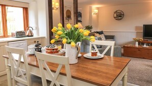 Uma área de refeições com mesa de madeira, cadeiras brancas, um vaso de flores amarelas e um bolo sobre a mesa.