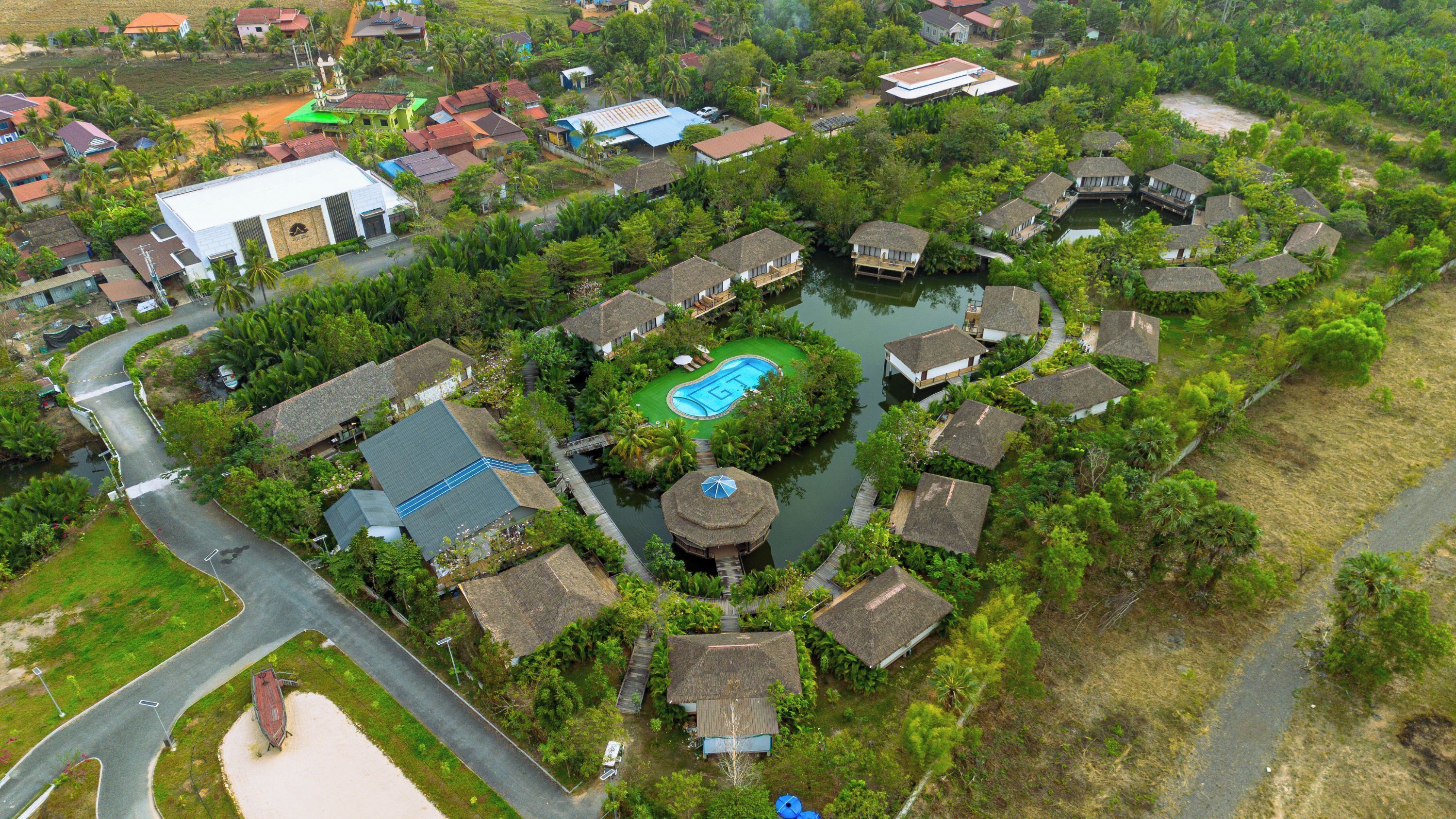 Vista aérea de um resort com piscina central, cercada por acomodações tipo bangalô, um edifício principal e uma estrada.