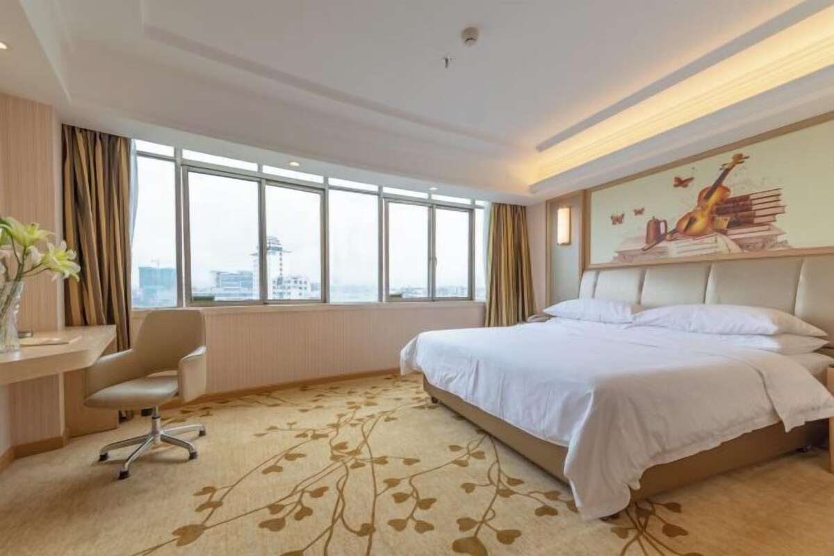 Quarto de hotel com uma cama grande, uma escrivaninha com cadeira, vista para a paisagem urbana através de janelas amplas e um mural na parede.