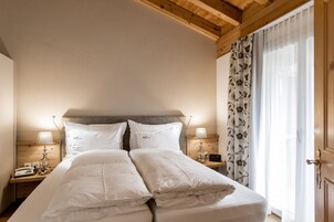Quarto de hotel com teto de madeira, cama com roupa de cama branca, duas luminárias de cabeceira e uma janela com cortinas florais.