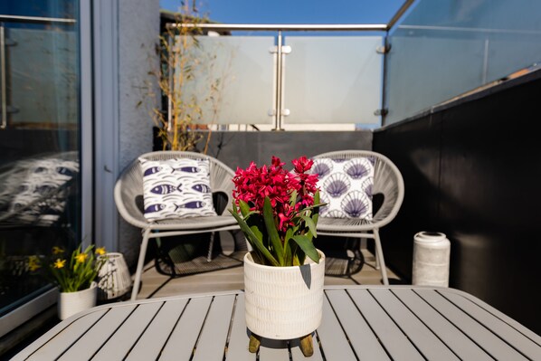 Uma pequena área de pátio com uma mesa branca, duas cadeiras de vime e um vaso branco com flores cor-de-rosa.