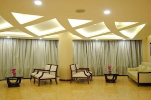 Um hall com teto alto, iluminação embutida, uma coluna imponente e uma área de estar com cadeiras e um sofá.