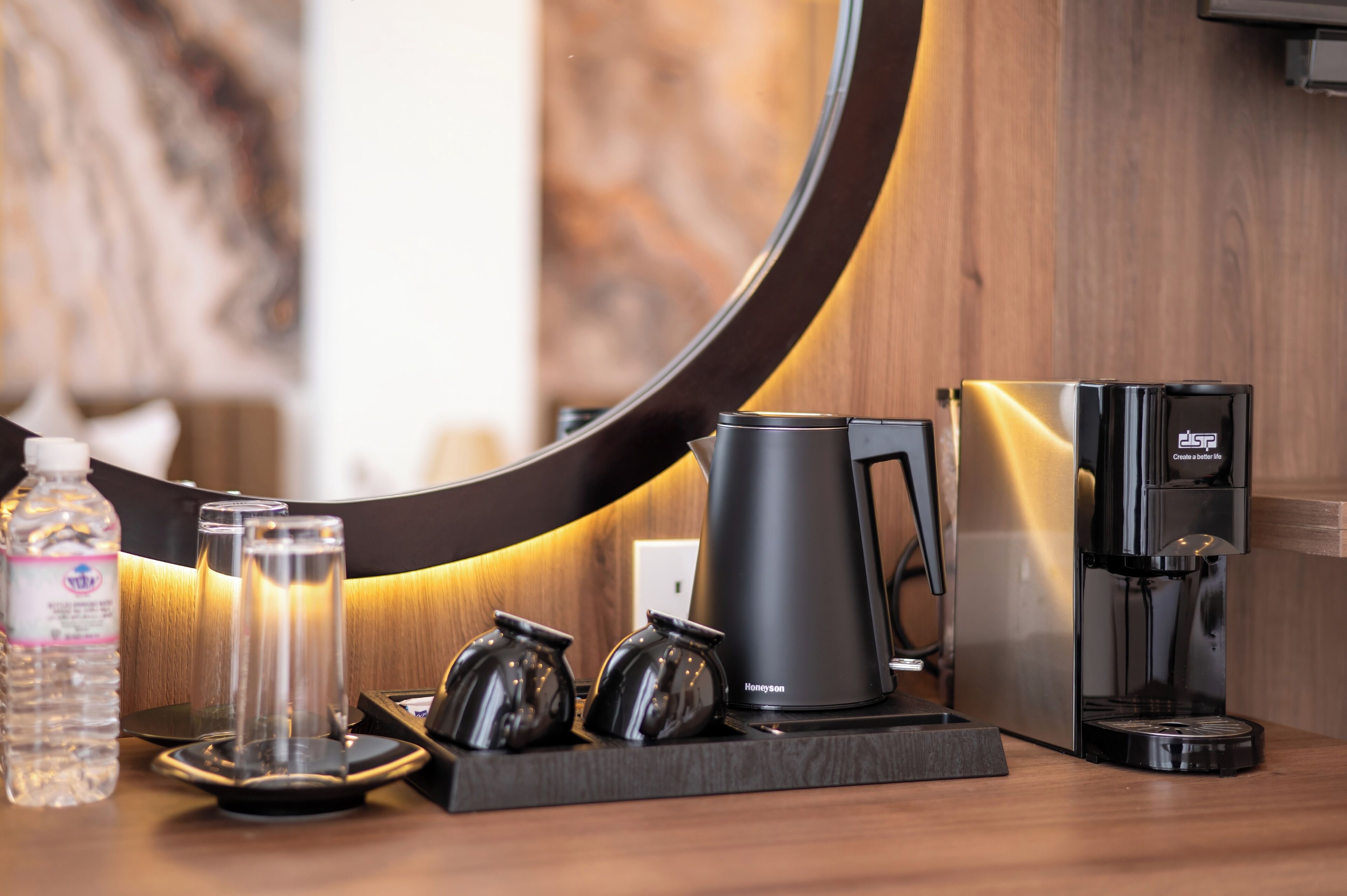 Quarto de hotel com parede de madeira, um espelho redondo, máquina de café preta, chaleira, garrafa de água e dois copos sobre uma bandeja.