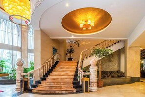 Uma imponente escadaria com corrimão, conduzindo a um andar superior, cercada por um amplo hall com um grande lustre circular.