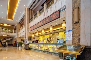 Um hall de hotel com um balcão de recepção de mármore, uma escadaria e um painel de informações digital.