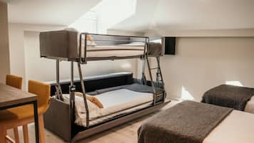 Uma cama beliche com escrivaninha e cadeiras, um claraboia e uma televisão na parede.