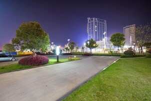 Rua urbana bem iluminada à noite, com um prédio alto ao fundo, uma estrada livre e áreas verdes.