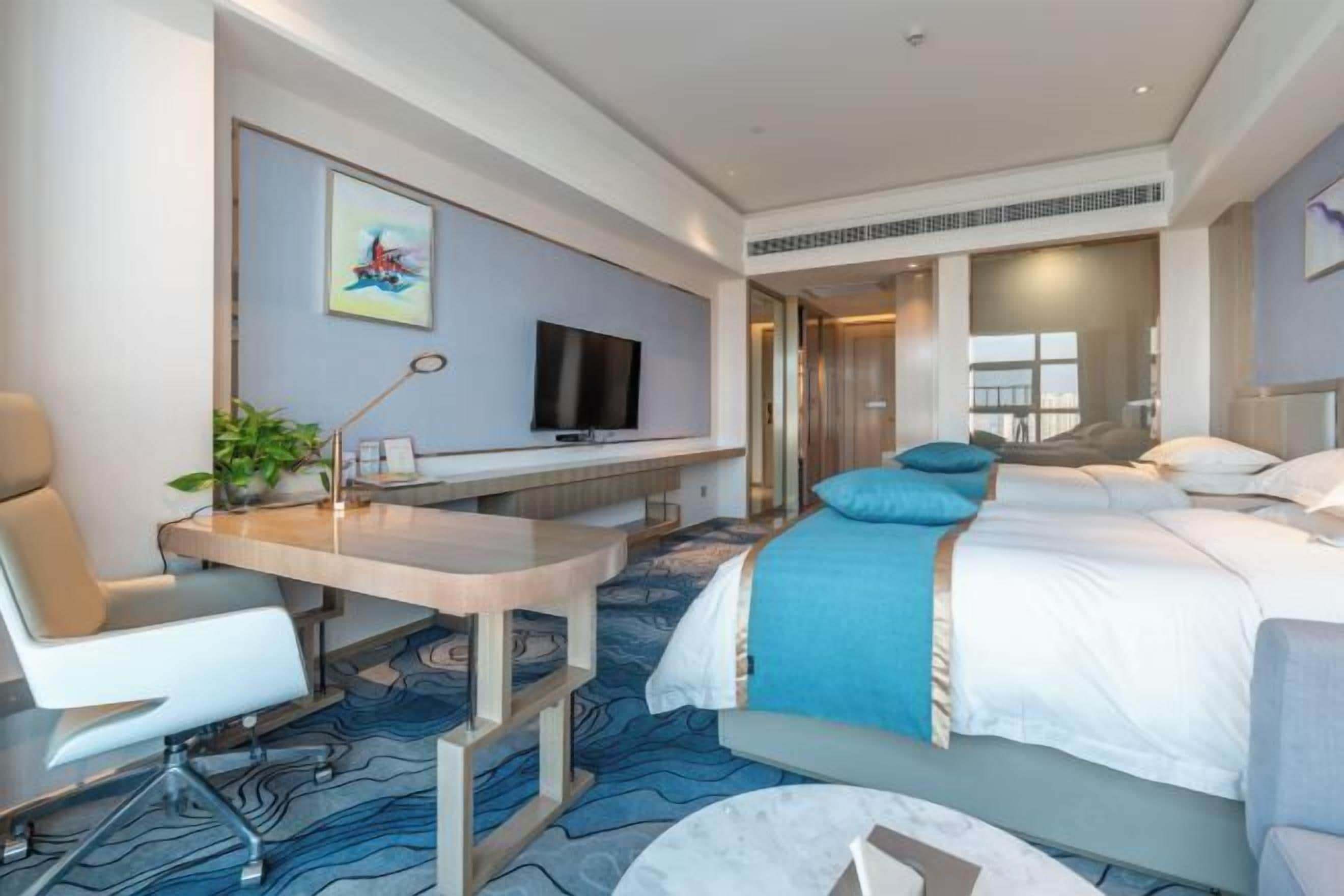 Quarto de hotel com uma cama, uma escrivaninha, uma cadeira, uma televisão e uma planta.