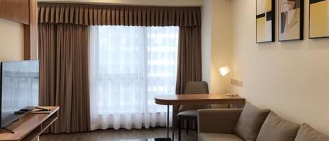 Quarto de hotel moderno com televisão de tela plana, mesa de centro de madeira, sofá, mesa de jantar com cadeiras e uma grande janela com cortinas.