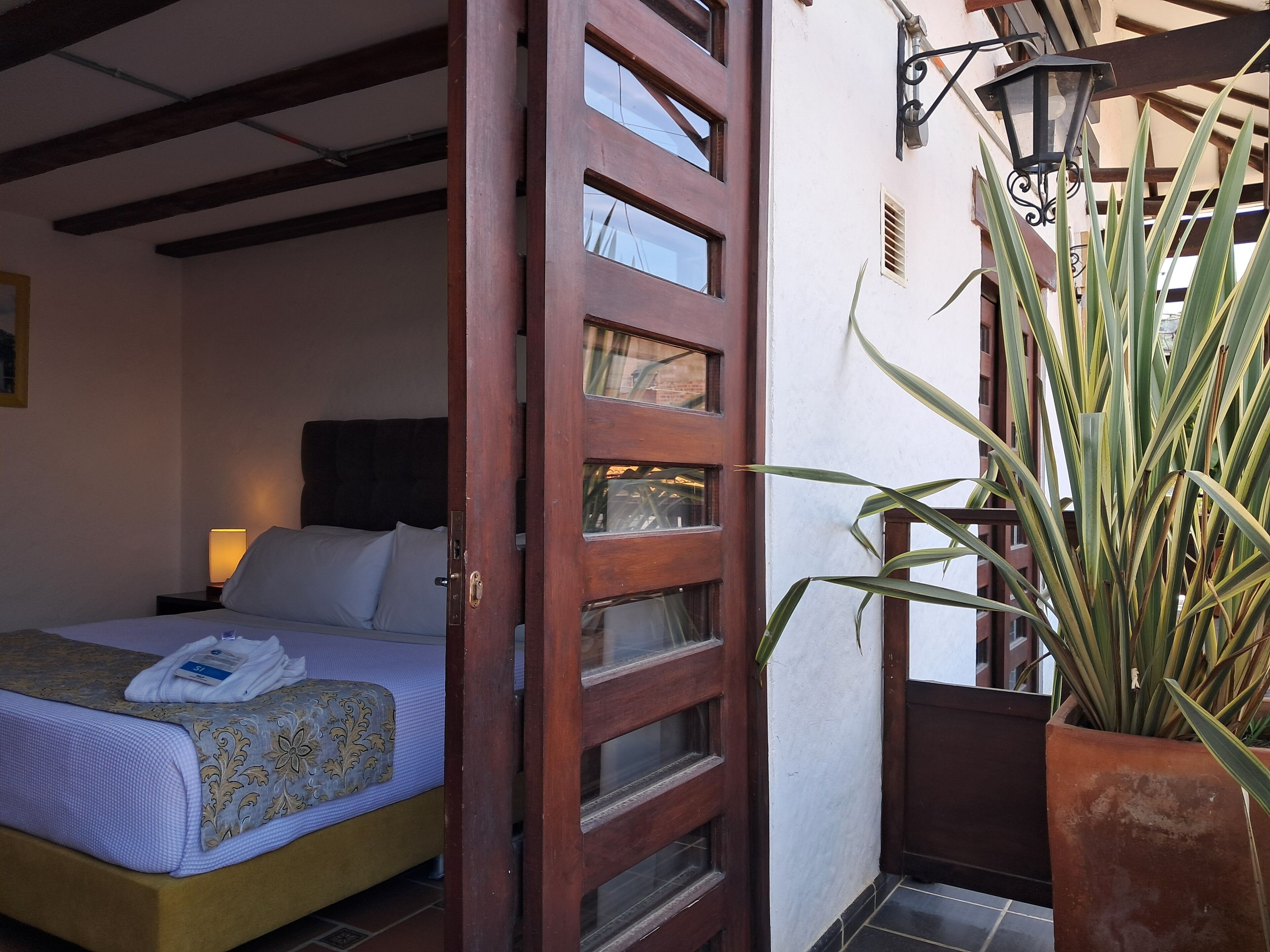 Quarto de hotel com cama, mesa de cabeceira, luminária e planta em vaso.