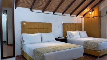 Quarto com duas camas, cada uma com cabeceira de madeira, roupa de cama branca e colcha estampada. Um criado-mudo com uma luminária e um telefone estão em uma das camas. O quarto possui piso de azulejos e teto inclinado com vigas de madeira.