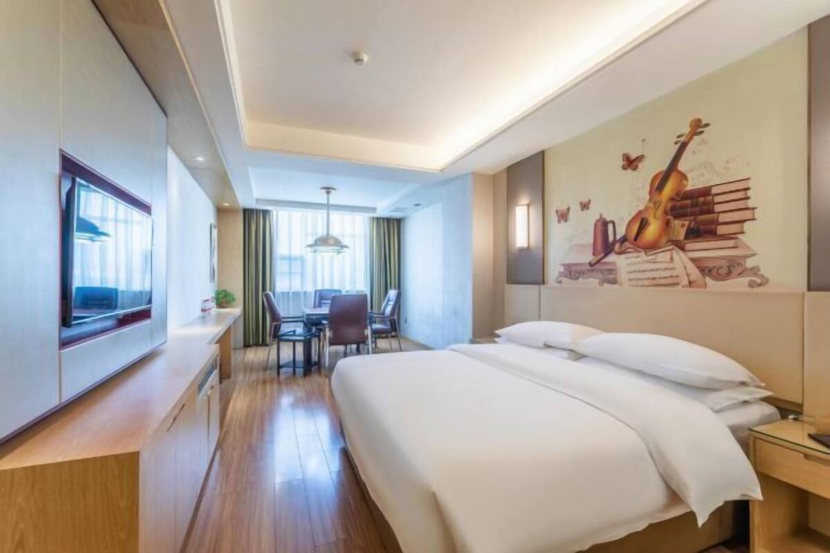 Quarto de hotel com cama, escrivaninha, cadeira, televisão e parede com instrumento musical e livros.
