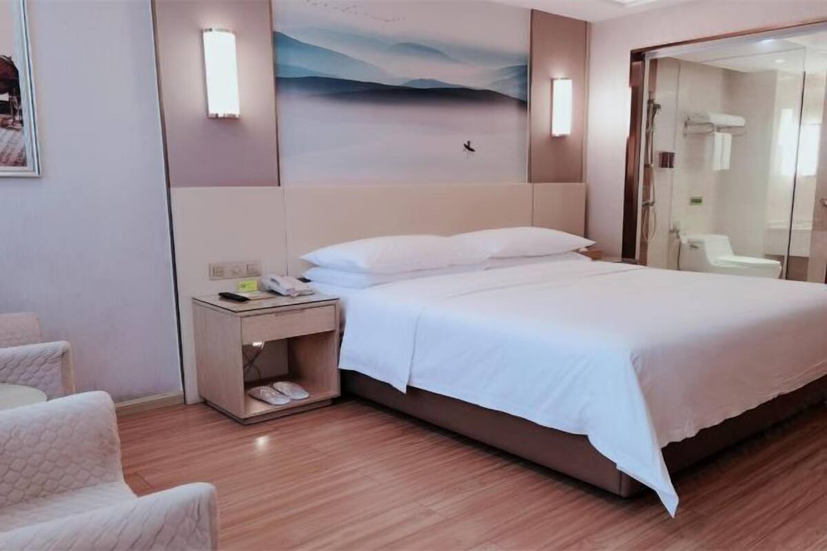 Quarto de hotel com uma cama grande, mesa de cabeceira com telefone, criadeira e área de chuveiro com divisória de vidro.