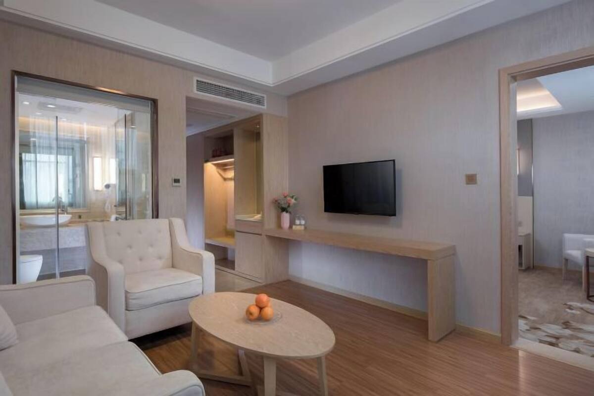 Quarto de hotel moderno com sofá, mesa redonda com frutas, TV fixada na parede e um banco de madeira.