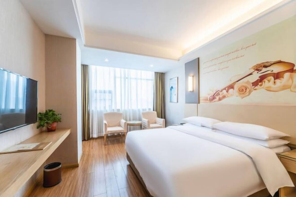 Quarto de hotel com uma cama, uma mesa de trabalho com televisão, uma cadeira, uma mesinha e uma planta.