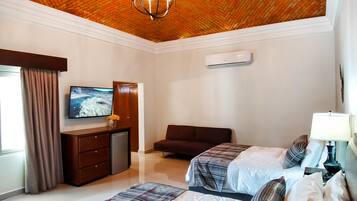Quarto de hotel com teto em padrão de tijolos, um lustre, uma TV, um sofá, duas camas com almofadas e uma mesa de cabeceira com abajur.