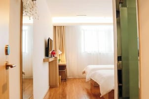 Quarto de hotel com piso de madeira, uma mesa com televisão, uma cama e uma janela com cortinas.
