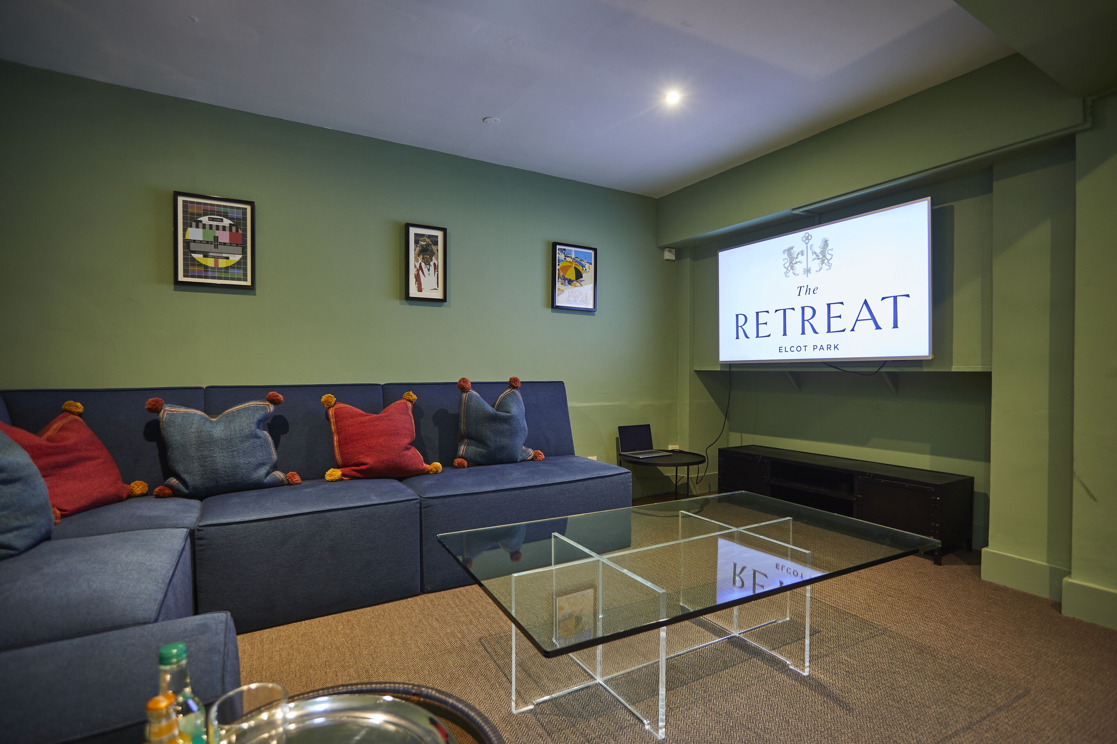 Uma sala de estar moderna com um sofá azul, uma mesa de centro de vidro e uma tela de projeção exibindo "The Retreat Elcot Park".