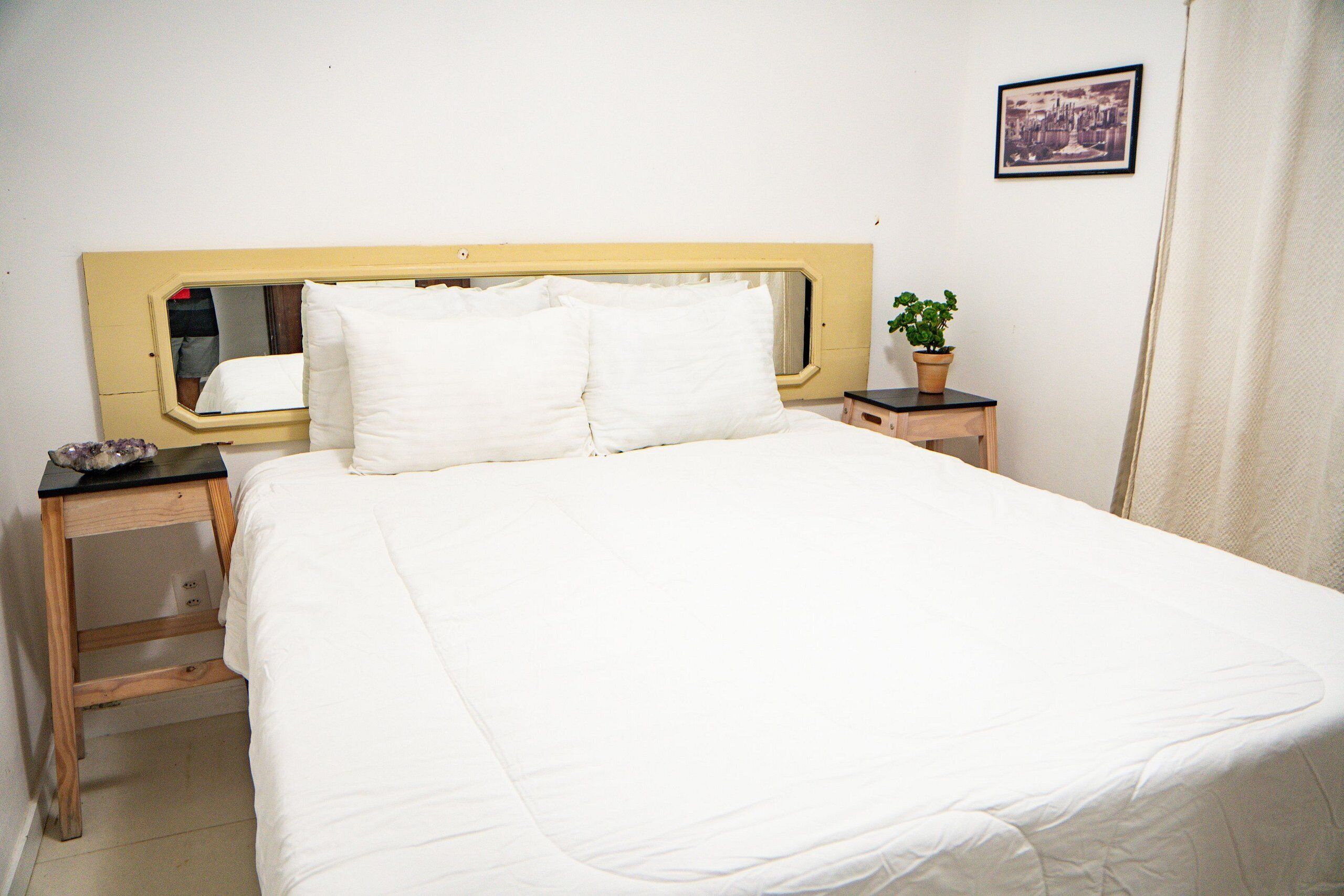 Uma cama com cabeceira de madeira, uma mesinha de cabeceira com uma planta e um quadro na parede.