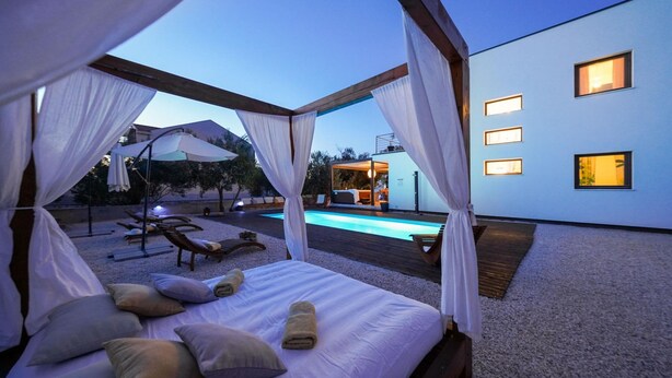 Uma cama com lençóis e almofadas brancos, um dossel e vista para uma piscina e um edifício ao fundo.