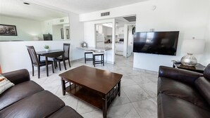 Uma sala de estar moderna com um sofá de couro marrom, uma mesa de centro de madeira, uma área de refeições com mesa e cadeiras, uma cozinha com geladeira e uma TV de tela plana fixada na parede.
