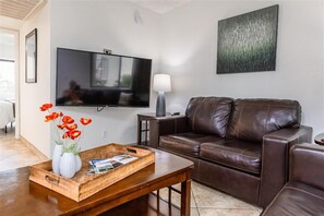 Uma sala de estar com um sofá de couro, uma televisão, um abajur e um tabuleiro com flores.