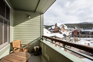 Varanda com cadeira e mesa de madeira, com vista para uma aldeia nevada, edifícios e montanhas.