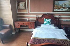Quarto de hotel com cama de madeira, mesa de cabeceira, escrivaninha e uma cadeira.
