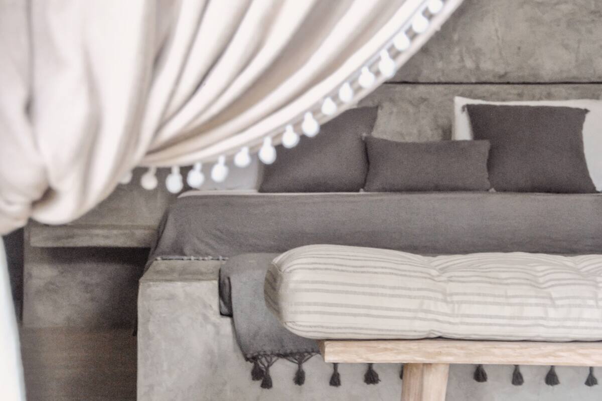Uma cama com roupa de cama cinza e almofada listrada, uma mesa de cabeceira de madeira e cortina com franjas.