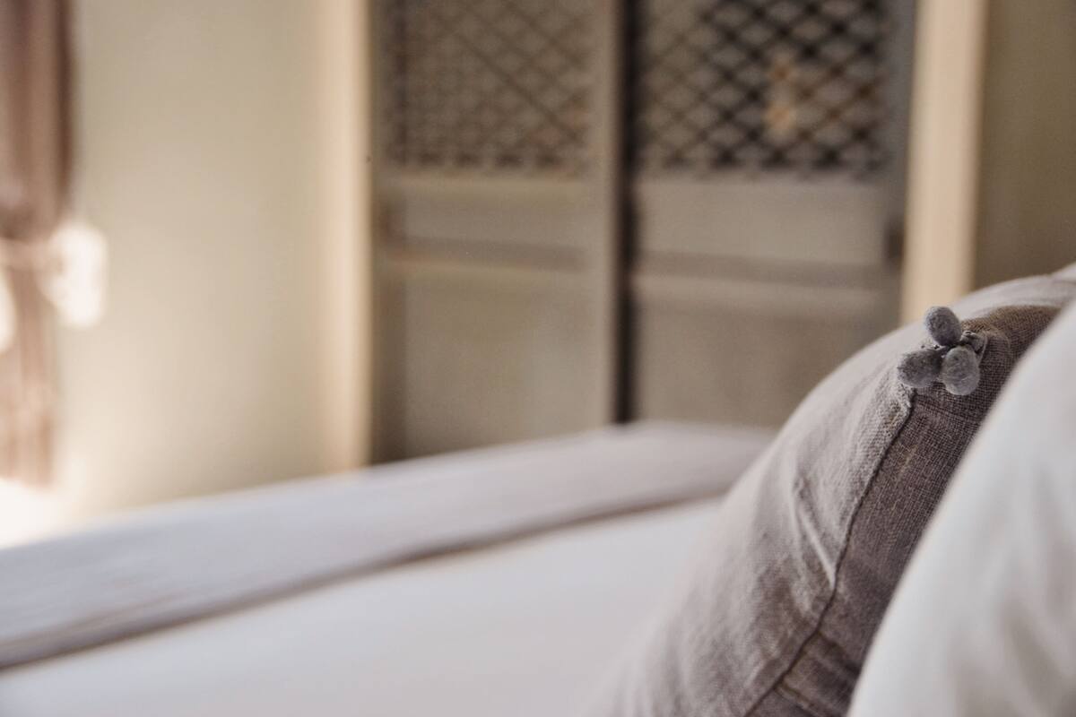 Um close-up de uma cama com colchão branco e almofada marrom com detalhe decorativo.
