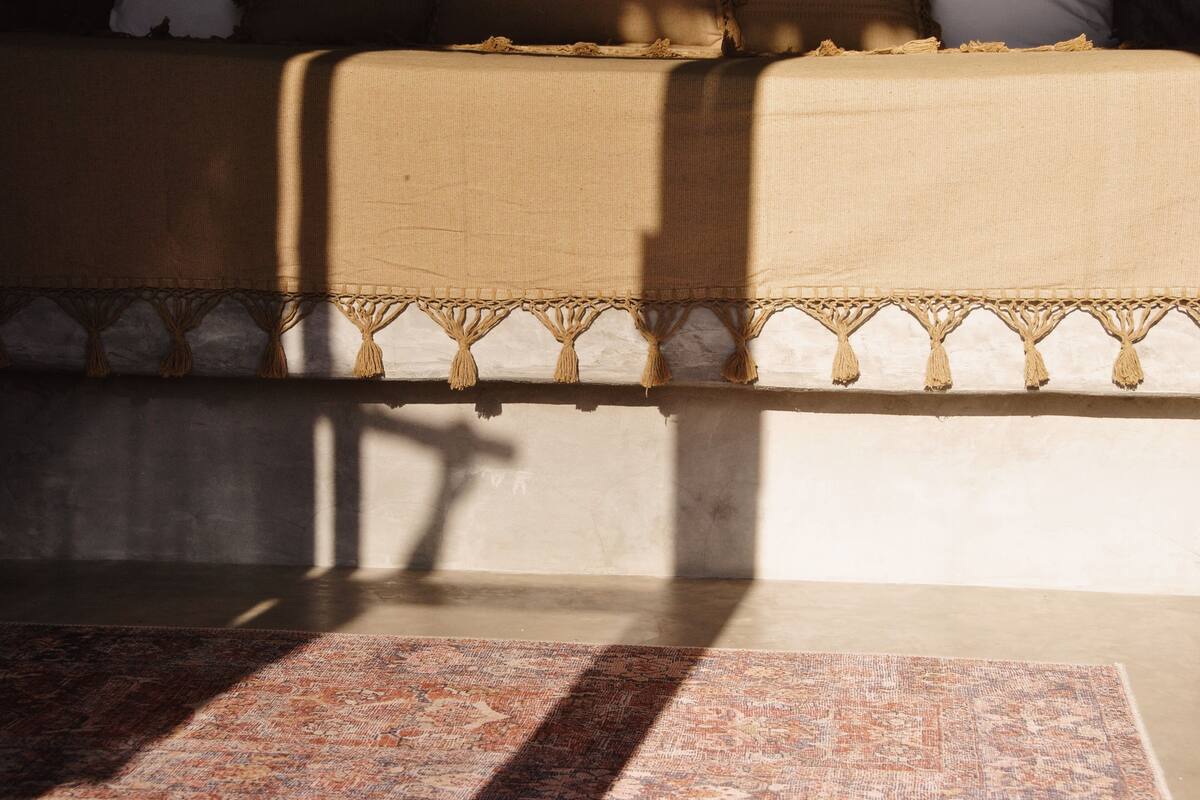 Quarto com sofá bege, tapete estampado e sombra projetada por uma janela.