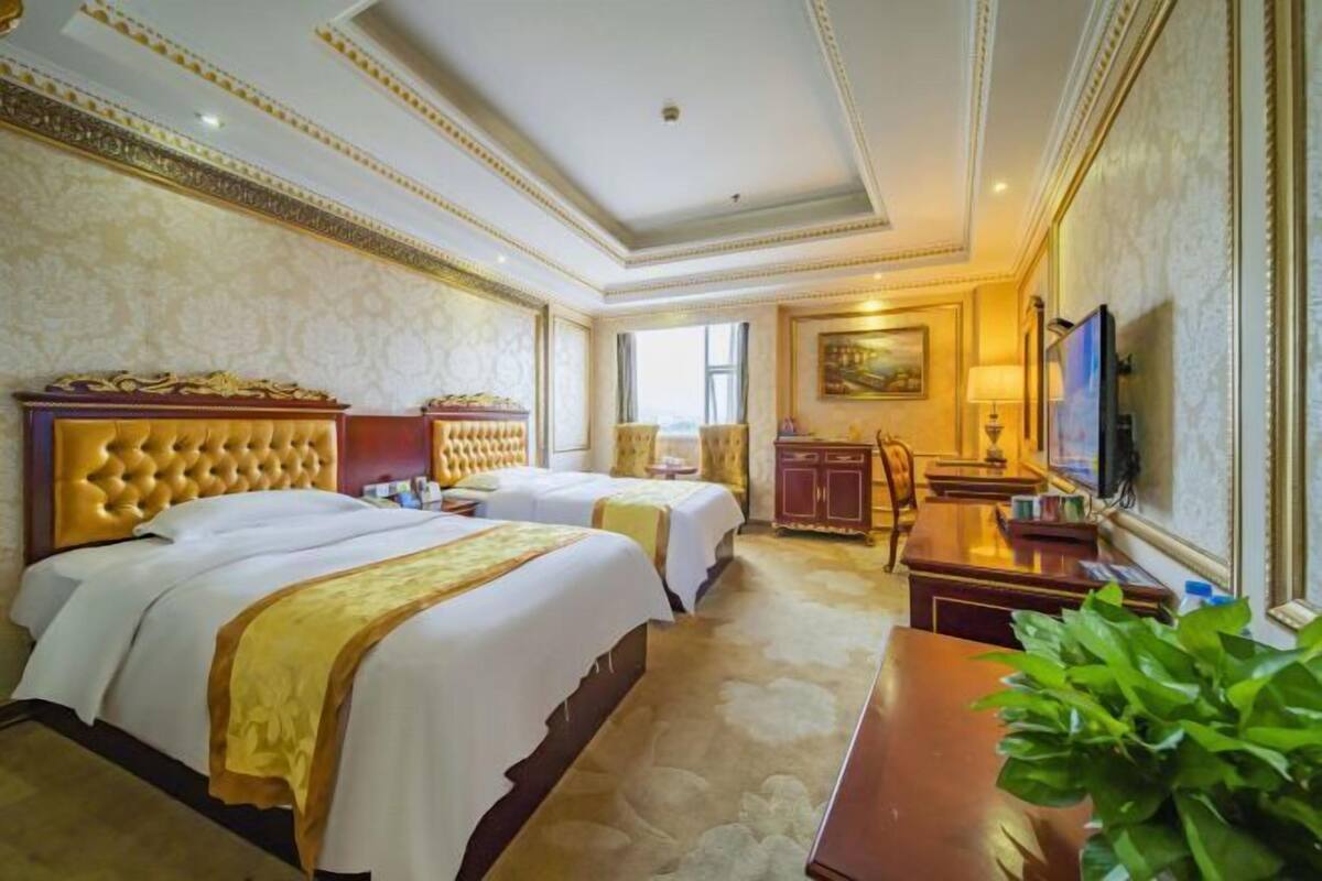 Quarto de hotel com duas camas, uma escrivaninha, uma televisão e um quadro na parede.