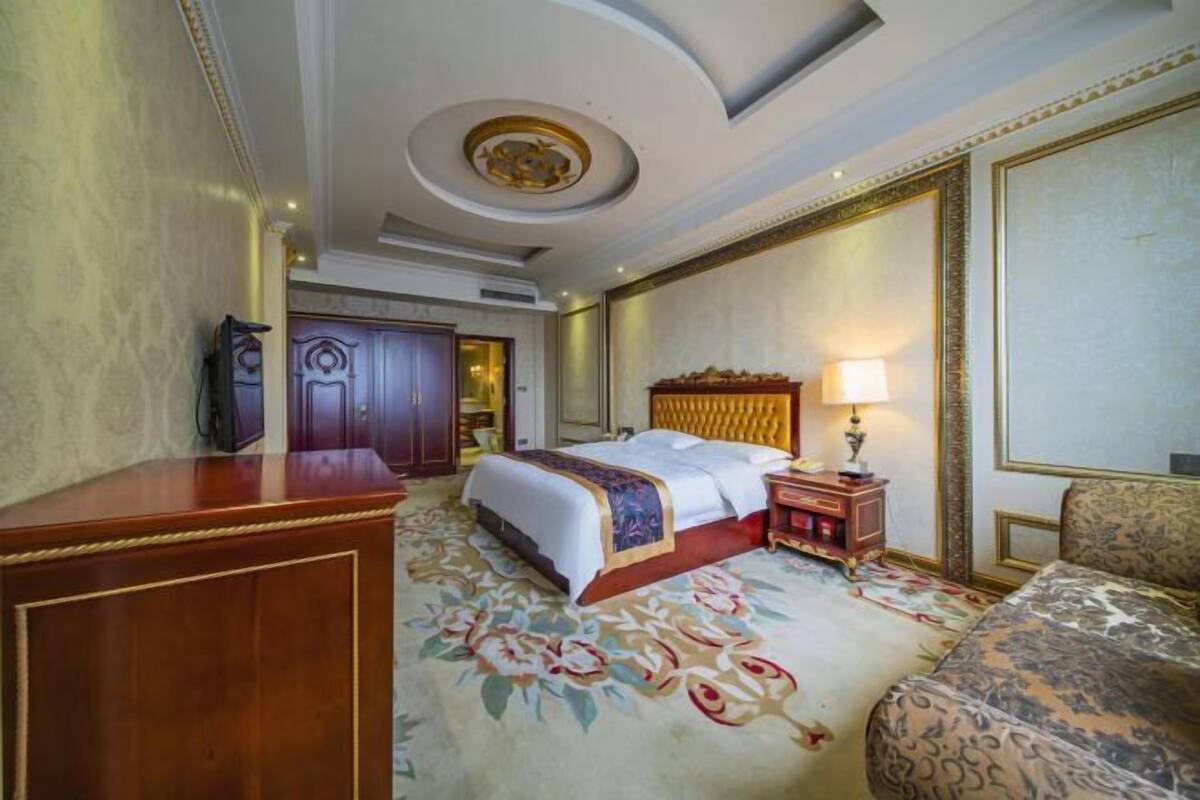 Quarto de hotel com uma cama grande, uma mesa de cabeceira de madeira, um sofá e teto decorativo.