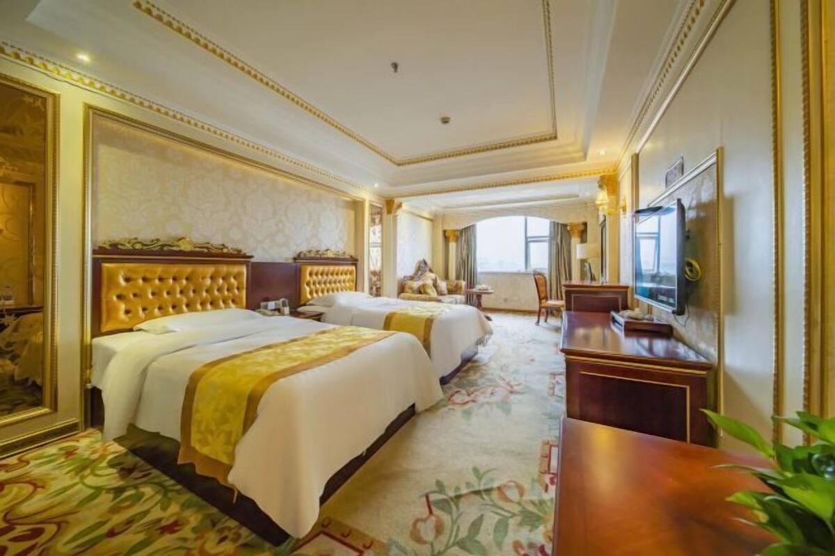 Quarto de hotel com duas camas, uma escrivaninha, uma televisão e uma janela grande.