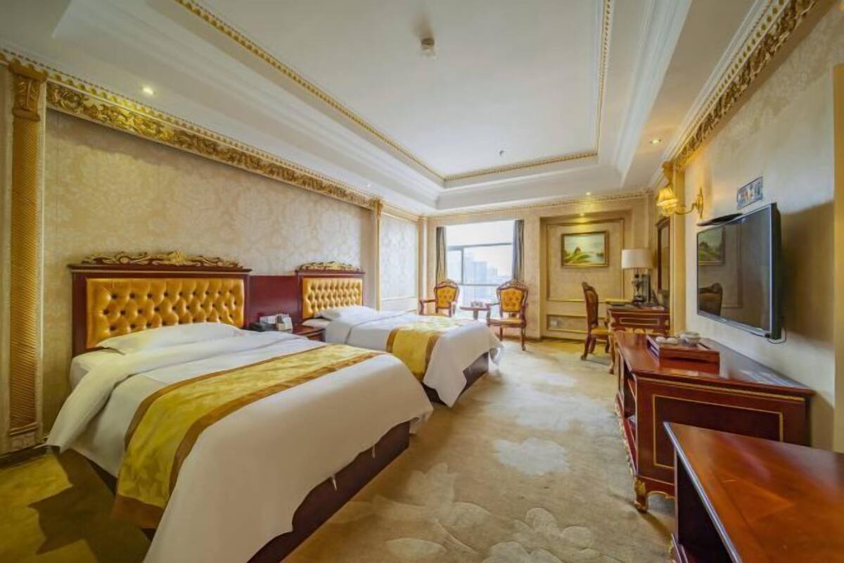 Quarto de hotel com duas camas, uma escrivaninha, uma cadeira, uma televisão e uma janela grande.
