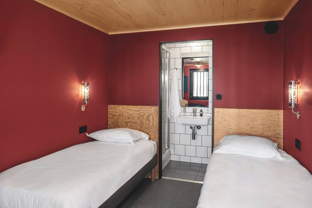 Quarto de hotel com duas camas de solteiro, teto de madeira, paredes vermelhas e banheiro com espelho e pia.