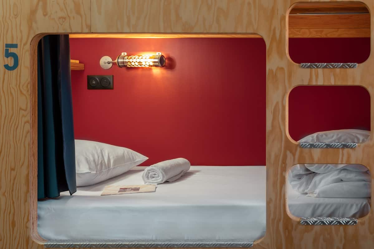 Quarto compacto com uma cama de solteiro, luminária fixada na parede e um espelho pequeno.