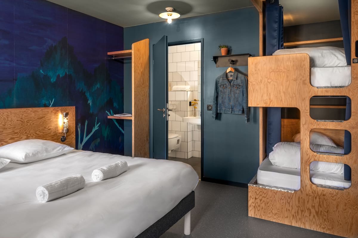 Quarto de hotel moderno com cama, beliche de madeira, banheiro com parede azul e um mural com tema de floresta na parede.