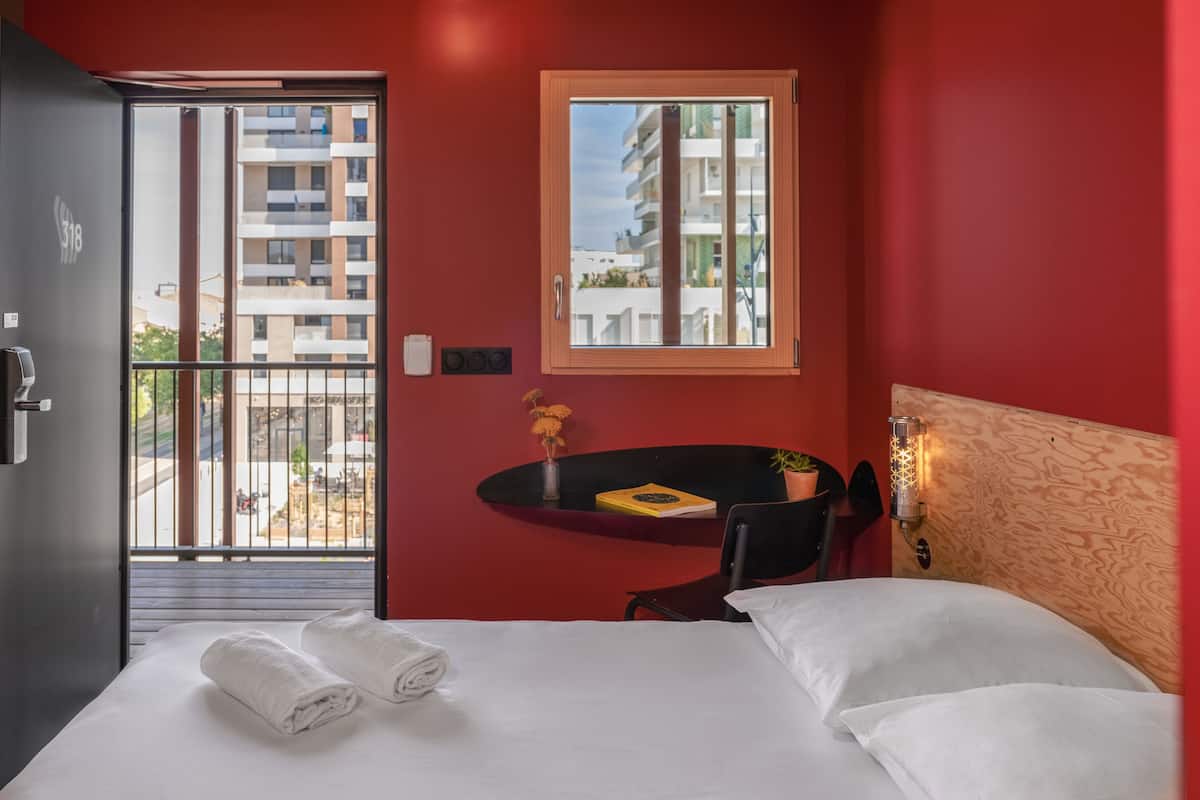 Quarto de hotel com cama, escrivaninha, cadeira, janela com vista para edifícios e porta para o exterior.