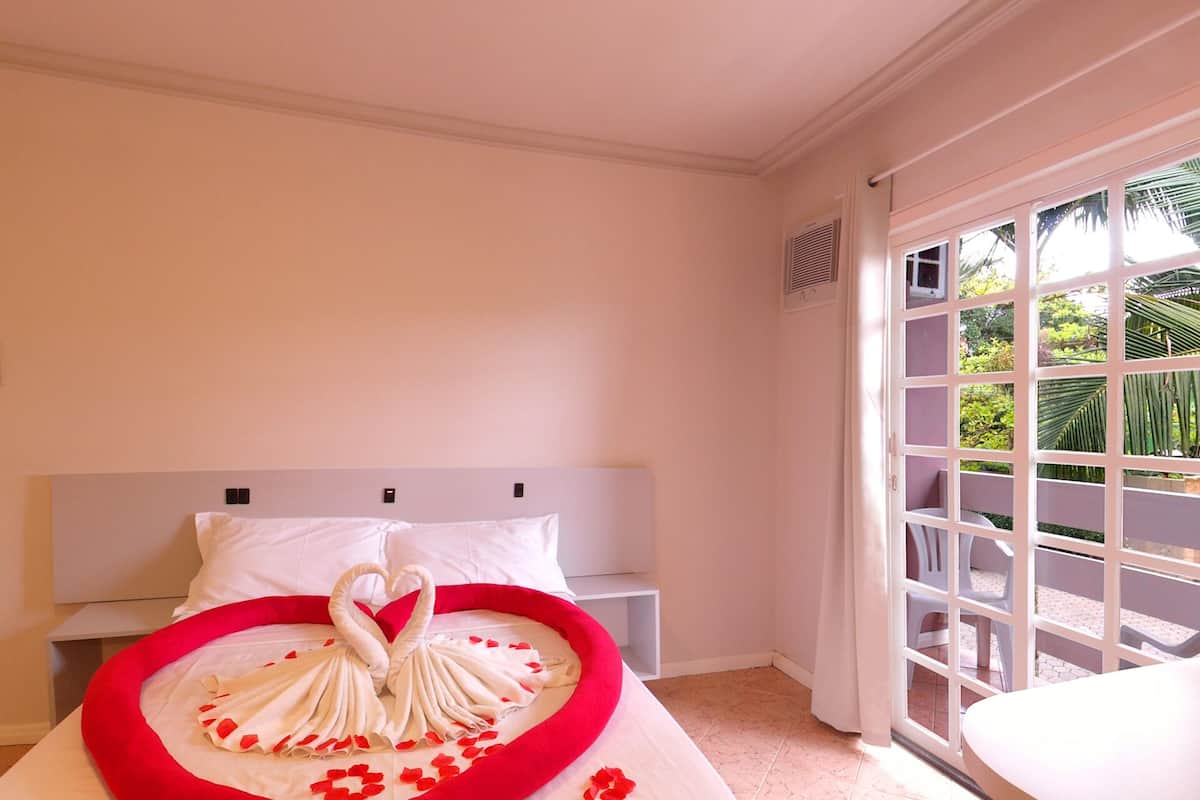 Quarto de hotel com cama, cabeceira branca, janela com vista para vegetação e mesa de cabeceira branca.
