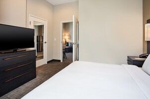 Quarto de hotel com uma cama grande, uma TV em uma cômoda, uma escrivaninha com luminária e uma porta para o banheiro.