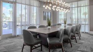 Uma sala de reuniões moderna com uma grande mesa, seis cadeiras cinzas, um lustre, janelas amplas com cortinas leves e um tapete estampado.