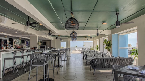 Um espaço interno amplo com piso de azulejos hexagonais, diversos arranjos de assentos incluindo banquetas e mesas, ventiladores de teto e luminárias pendentes.
