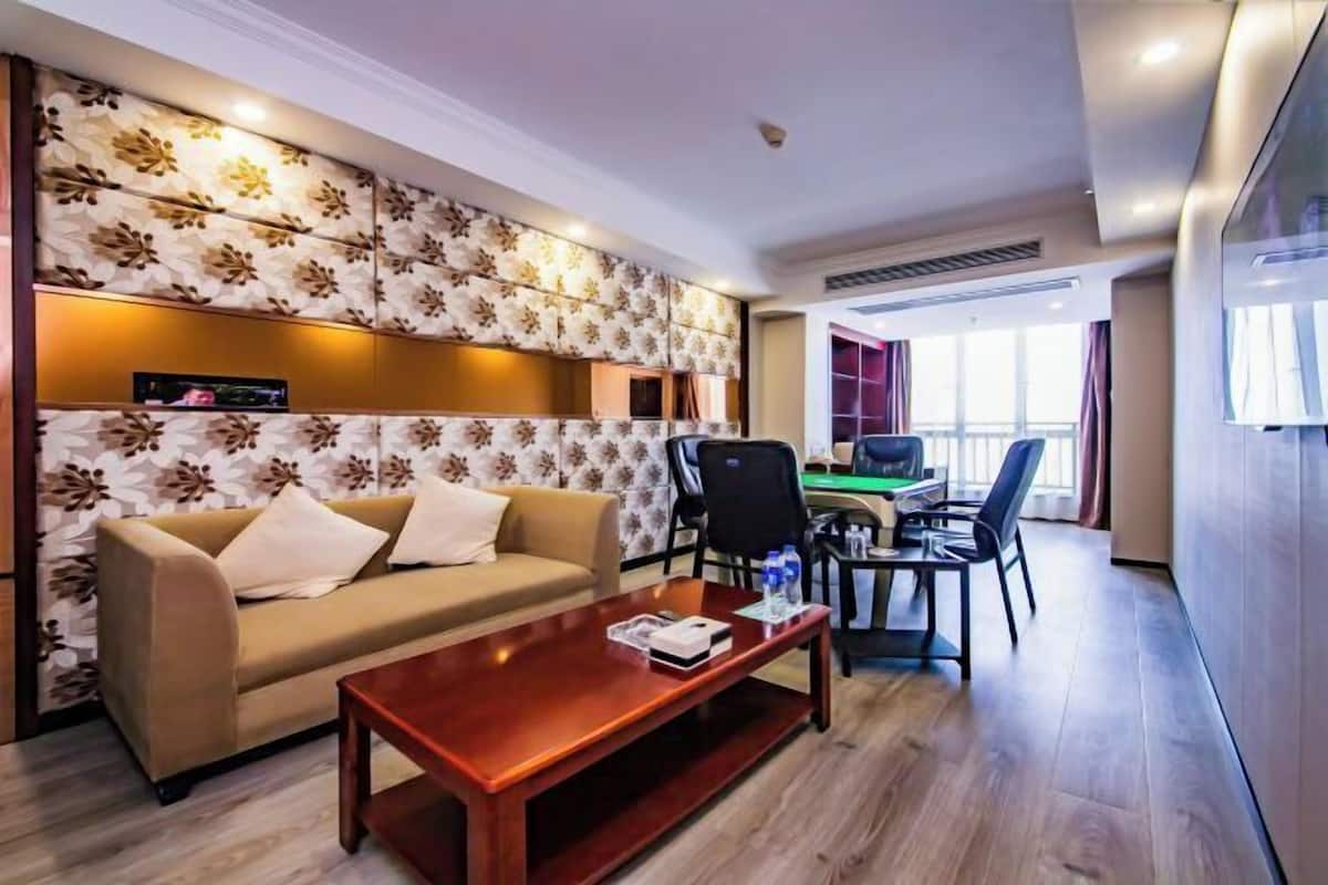 Quarto de hotel com sofá, mesa de centro de madeira, escrivaninha com computador e janela grande.