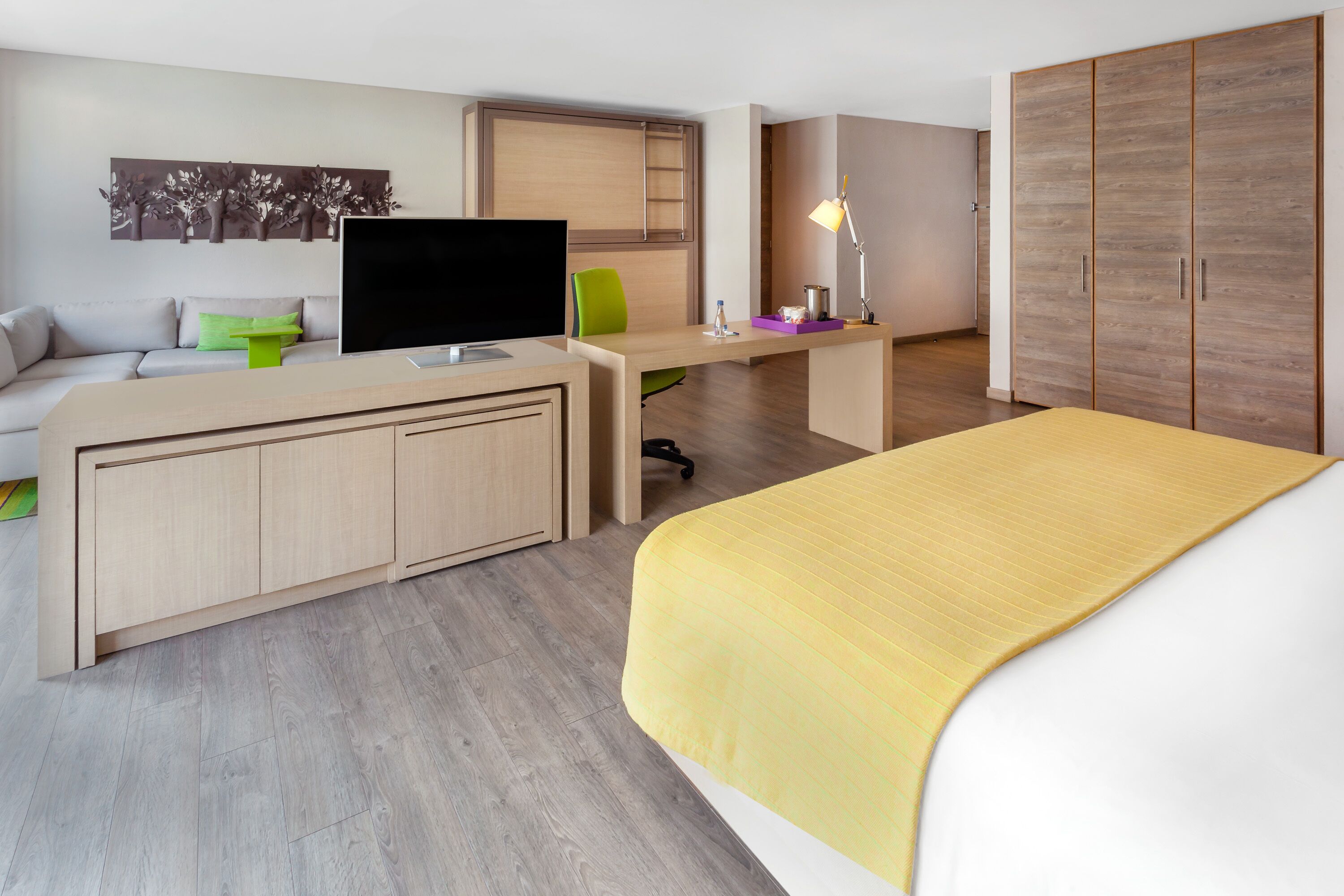Quarto de hotel moderno com cama, escrivaninha com computador, TV e guarda-roupa.